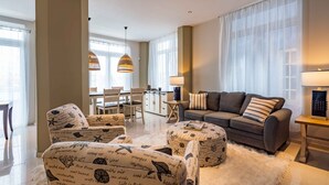 Uma sala de estar moderna com um sofá cinzento, poltronas estampadas, um tapete felpudo branco e uma área de jantar com mesa e cadeiras.