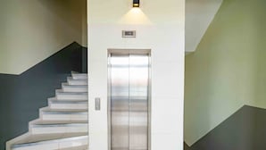 Um elevador moderno com visor digital, ladeado por uma escadaria de degraus claros e paredes em tons neutros.