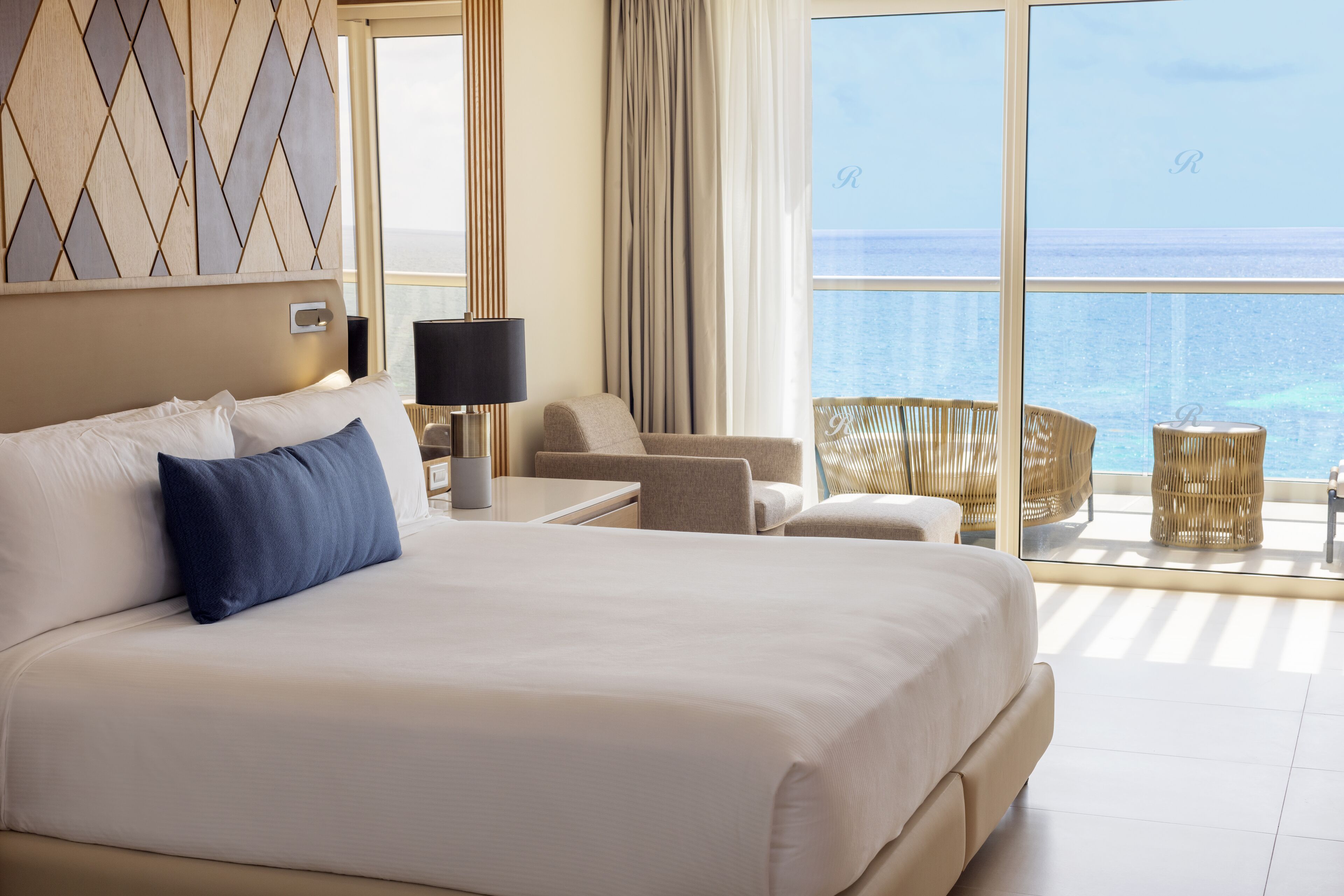 Uma cama grande com almofadas brancas e azuis, uma mesa de cabeceira com abajur e vista para o oceano através de grandes janelas.