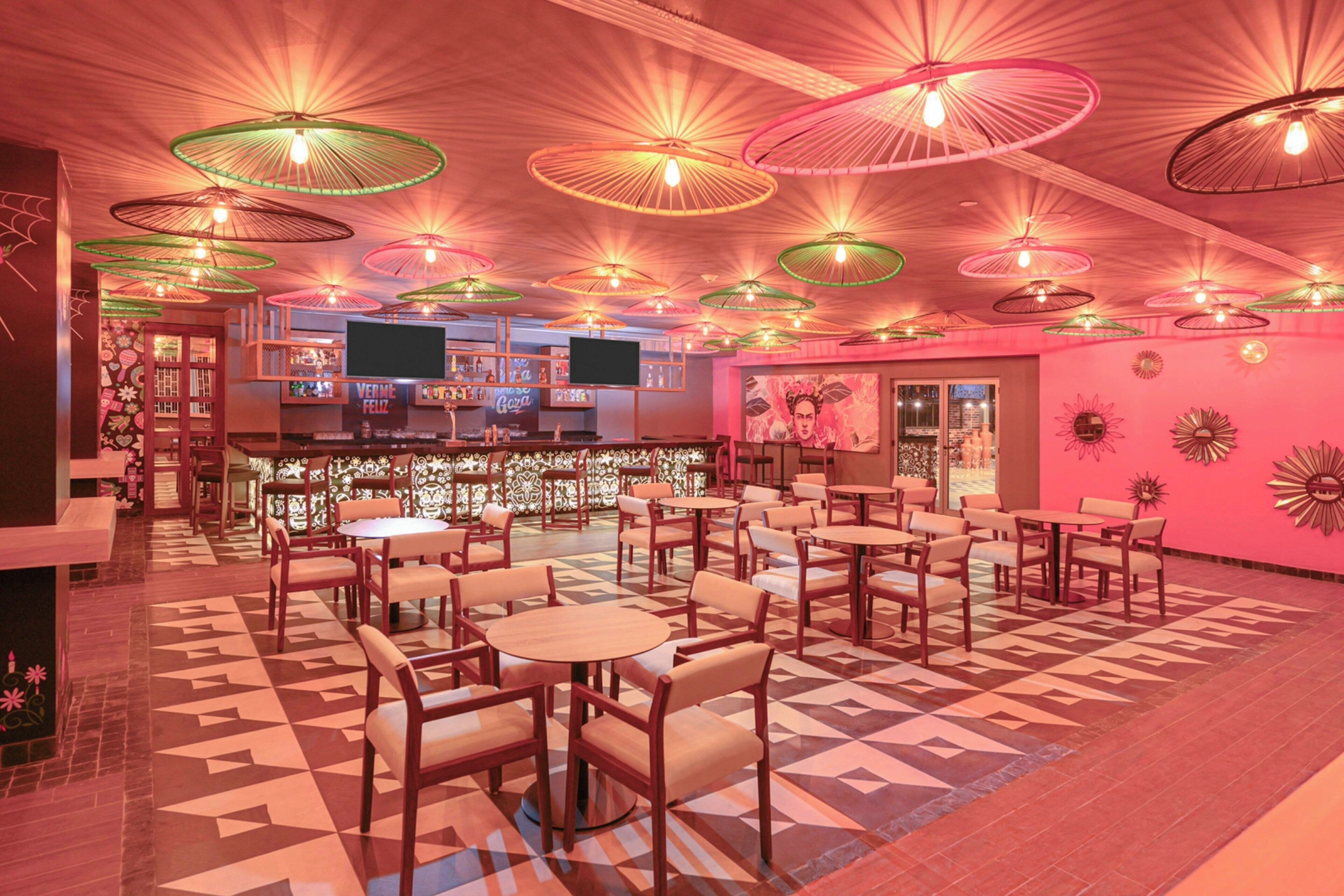 Uma área de bar com várias mesas e cadeiras, um balcão com uma televisão e um chão estampado.