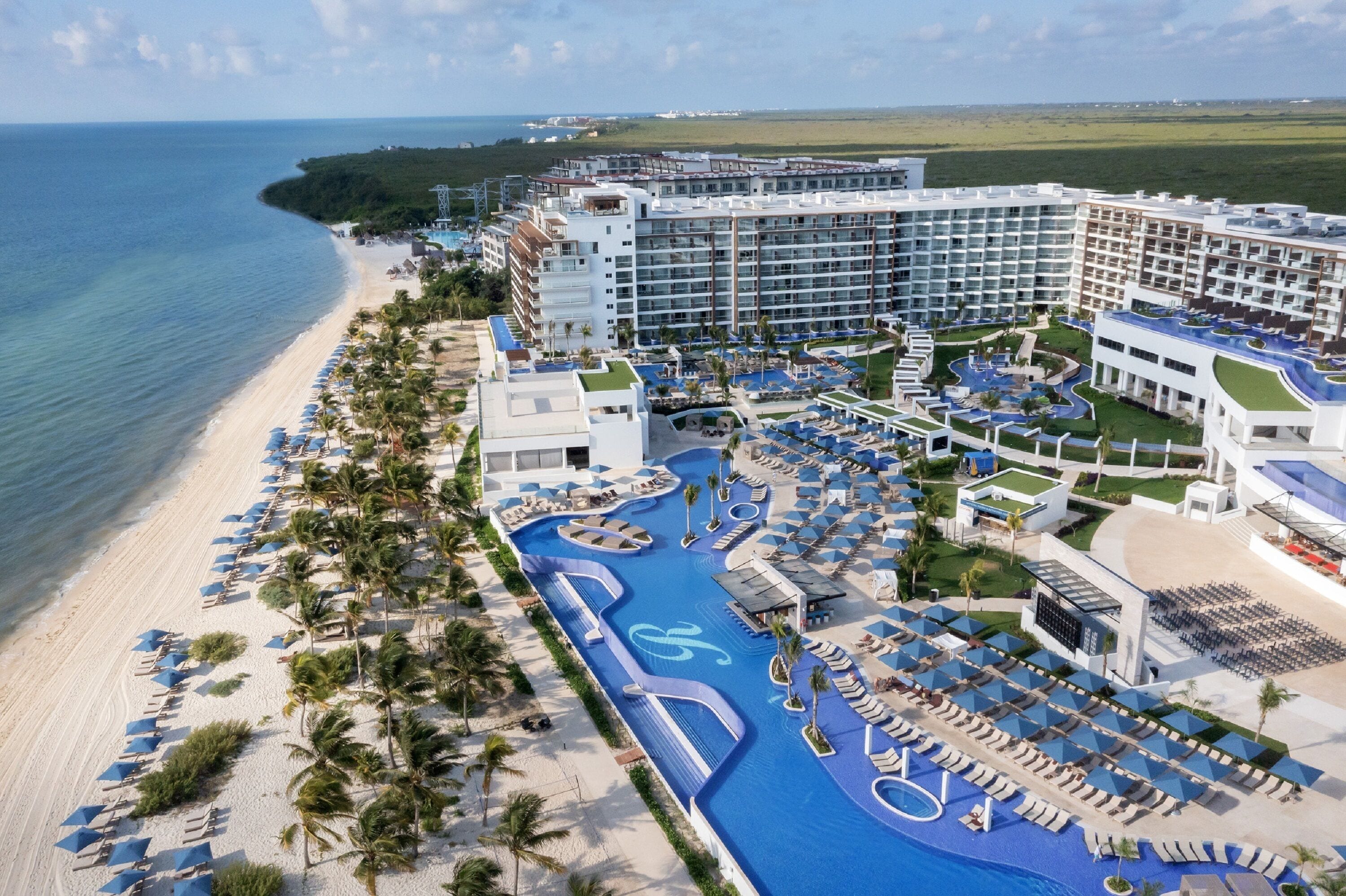 Um resort com uma piscina grande, várias áreas de lazer aquáticas e uma praia com guarda-sóis.