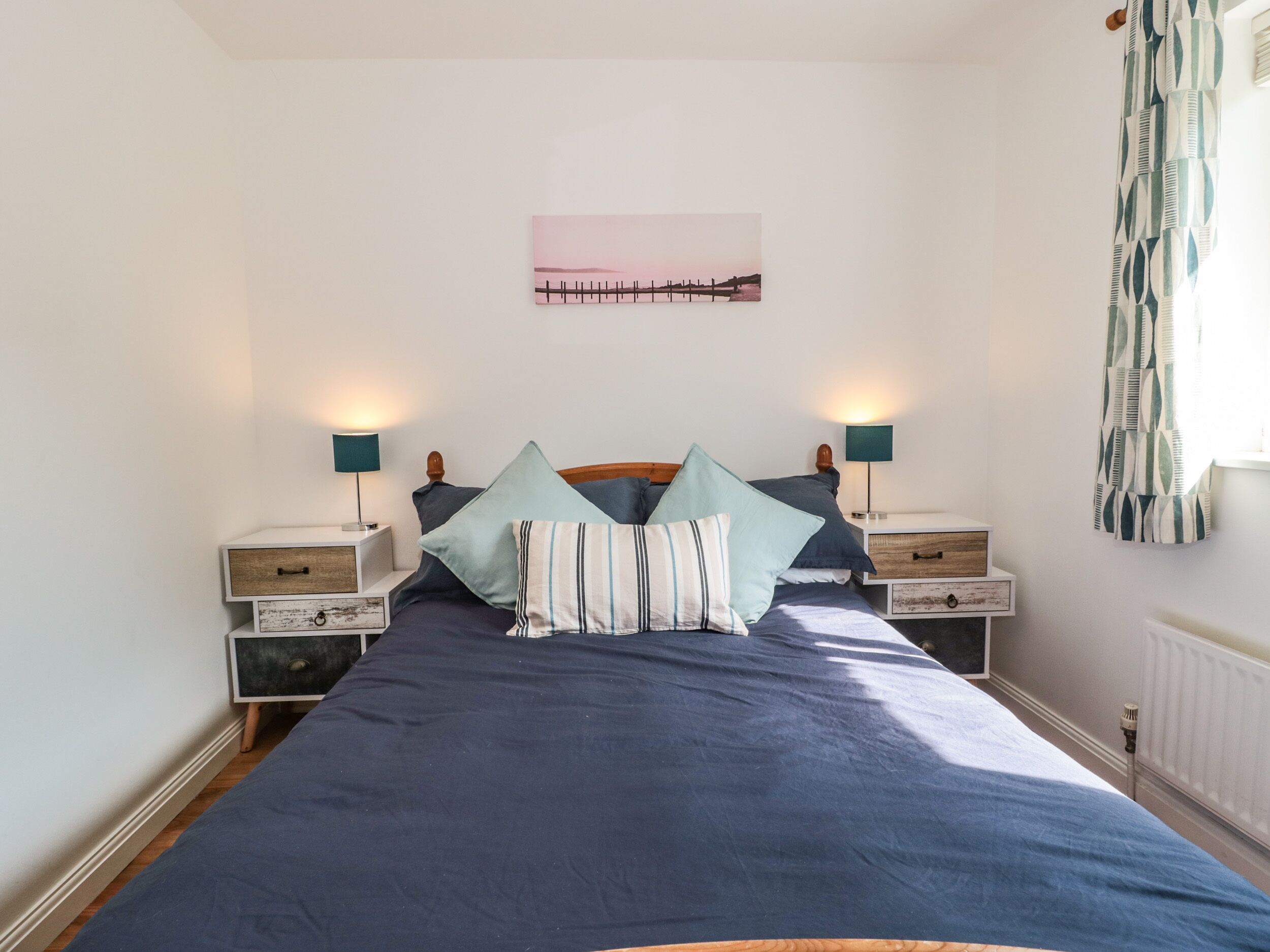 Quarto com uma cama grande, duas mesas de cabeceira com abajures, um quadro pendurado na parede e uma janela com cortinas.