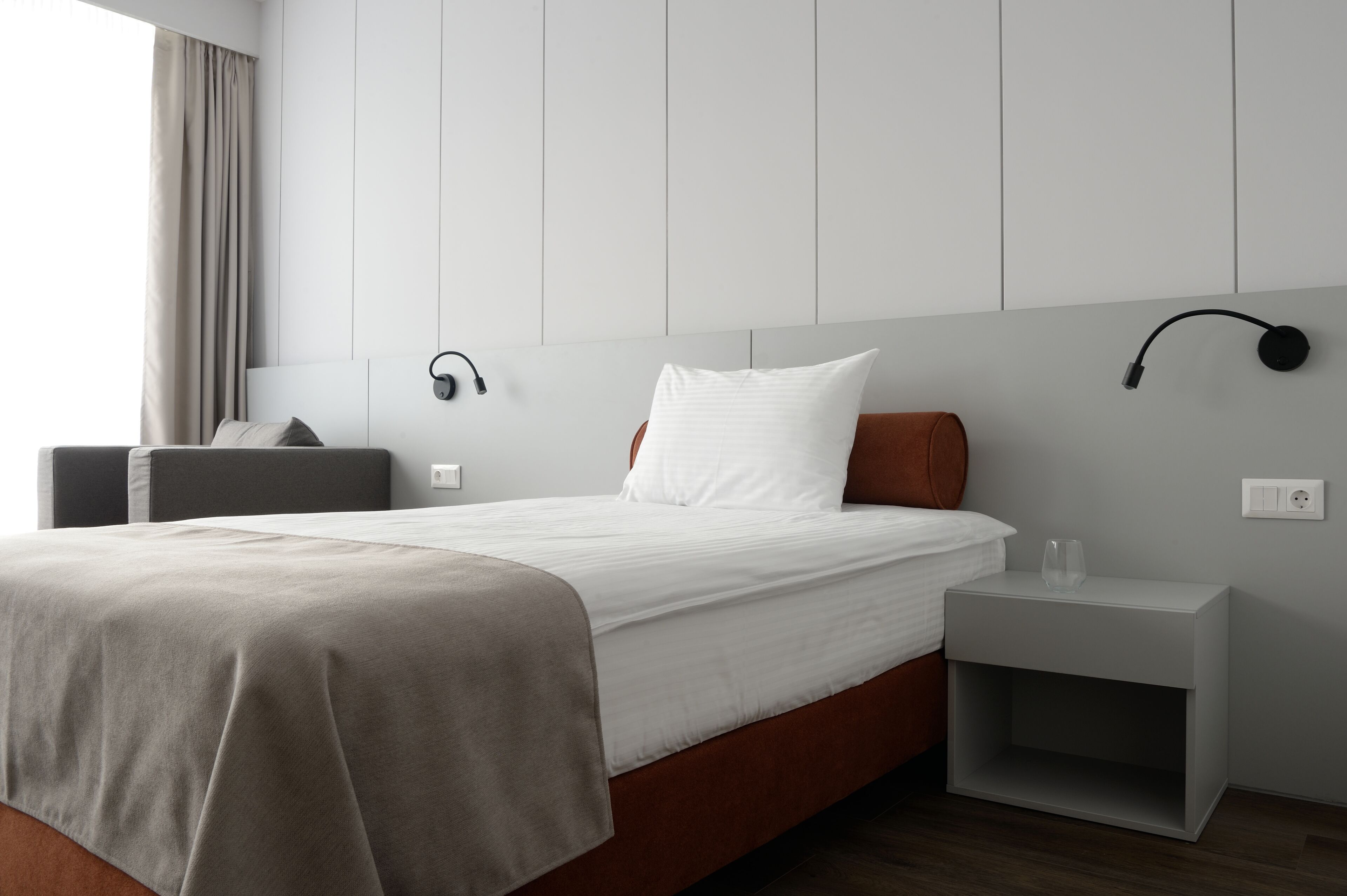 Quarto de hotel moderno com cama, mesa de cabeceira, cadeira e painel de cabeceira fixado na parede.