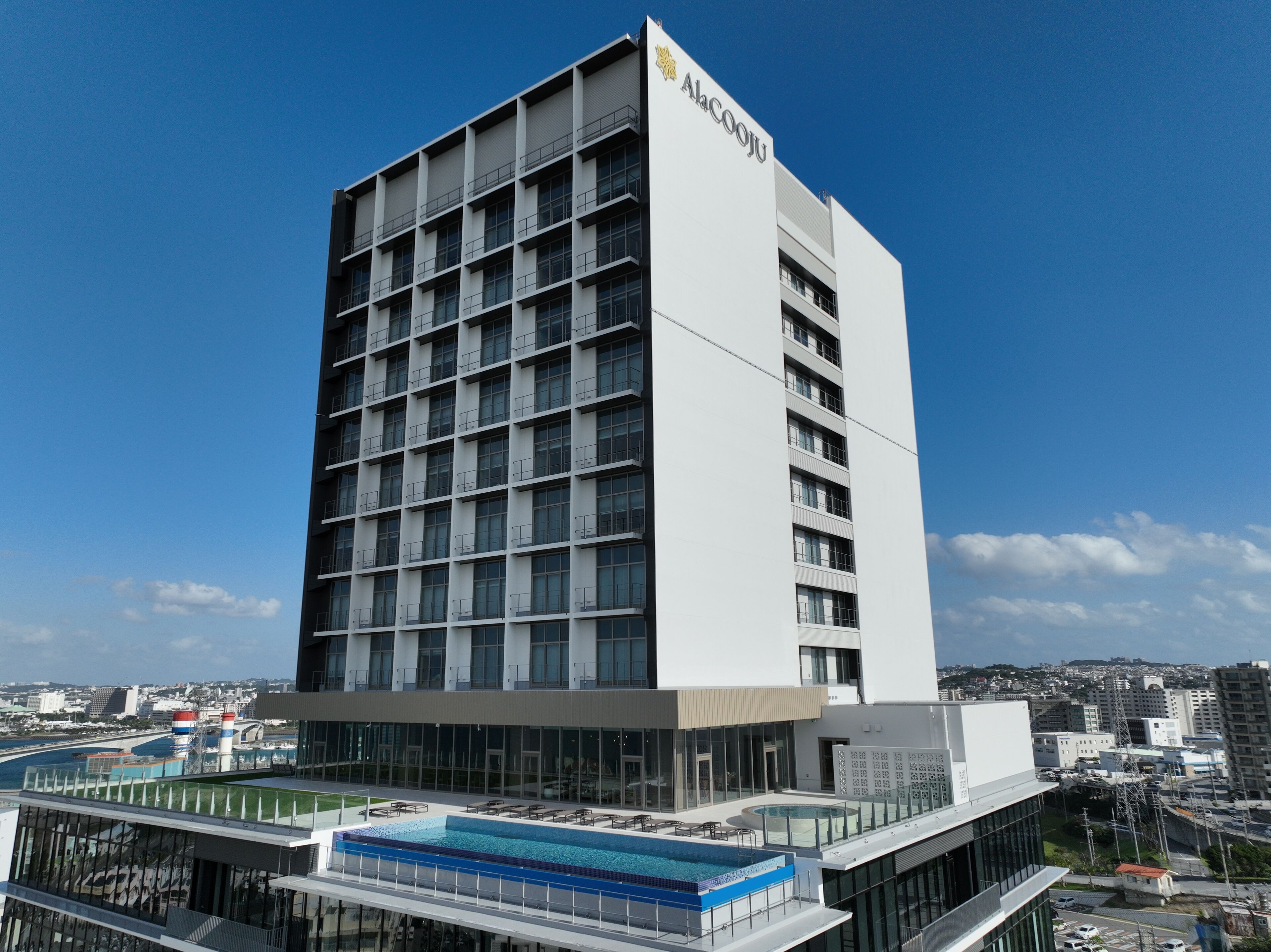 Um hotel moderno com uma placa 'Acooju' em destaque, apresentando uma área de piscina no terraço com espreguiçadeiras e uma piscina coberta por vidro.