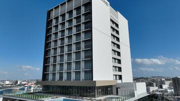 Um hotel moderno com uma placa 'Acooju' em destaque, apresentando uma área de piscina no terraço com espreguiçadeiras e uma piscina coberta por vidro.