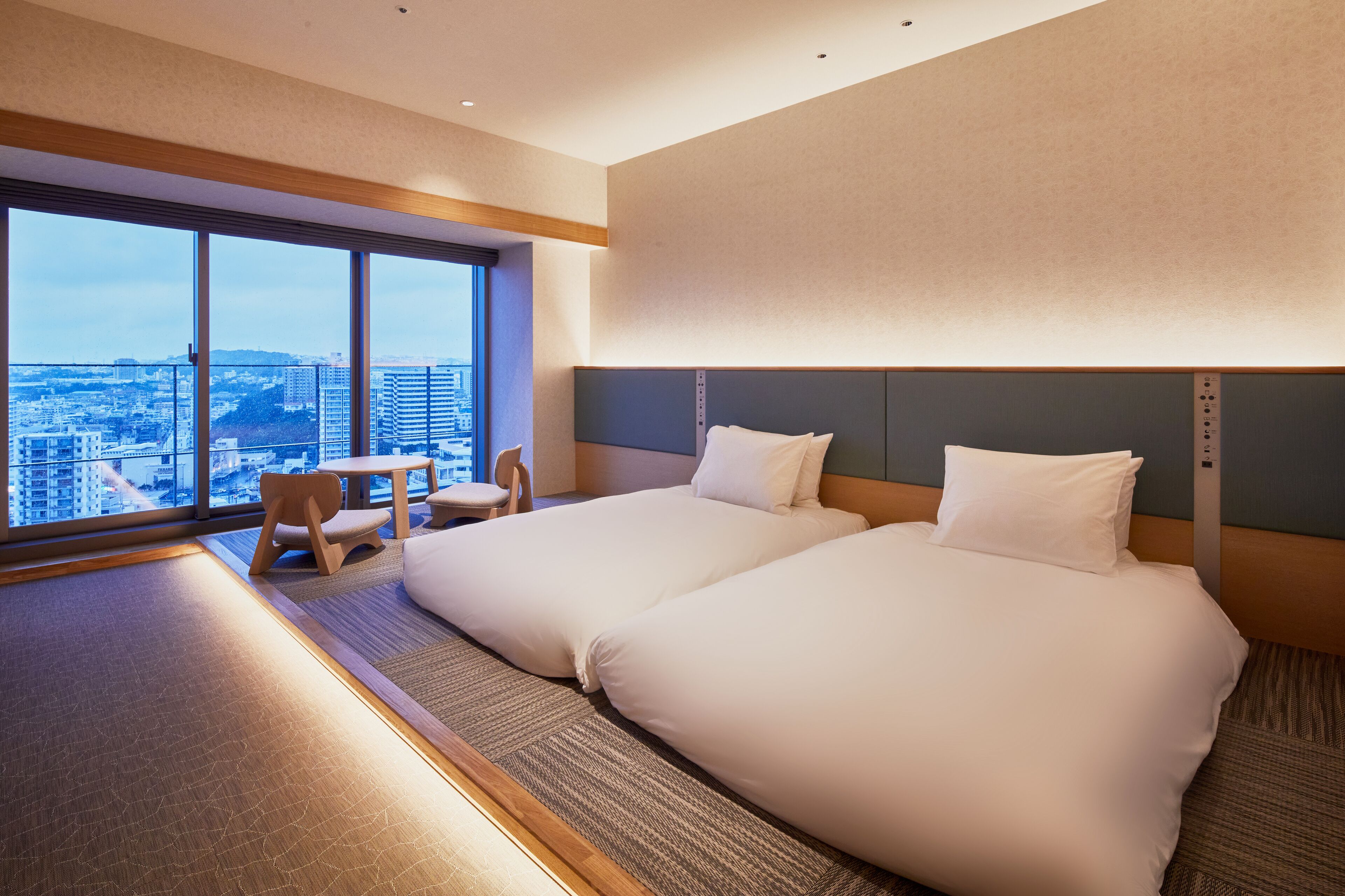 Quarto de hotel com duas camas, uma mesinha, uma cadeira e uma janela grande com vista para a cidade.