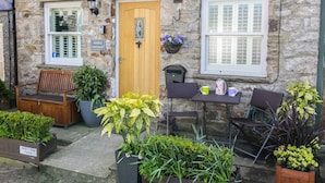 Um chalé de pedra com porta de madeira, duas janelas, um banco, uma mesa com cadeiras e plantas em vasos.