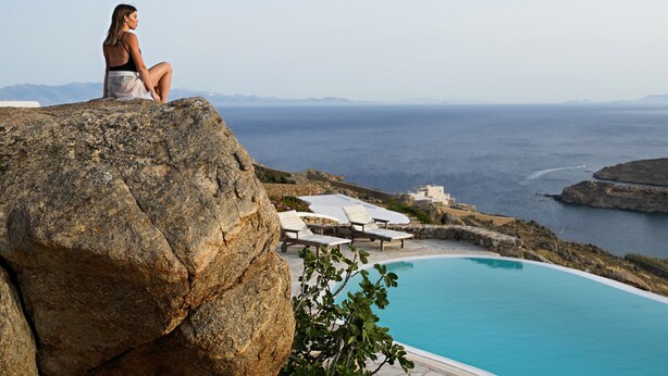Mulher sentada em uma rocha, com vista para uma piscina e o mar, além de montanhas ao longe.