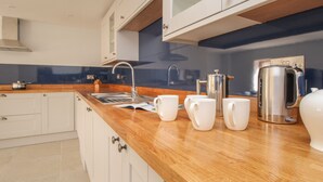 Cozinha moderna com armários brancos, bancada de madeira, cafeteira de aço inoxidável e quatro canecas brancas.