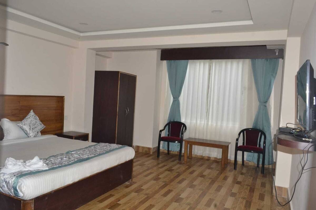 Quarto de hotel com cama, guarda-roupa, duas cadeiras, uma mesinha, televisão e janela com cortinas.