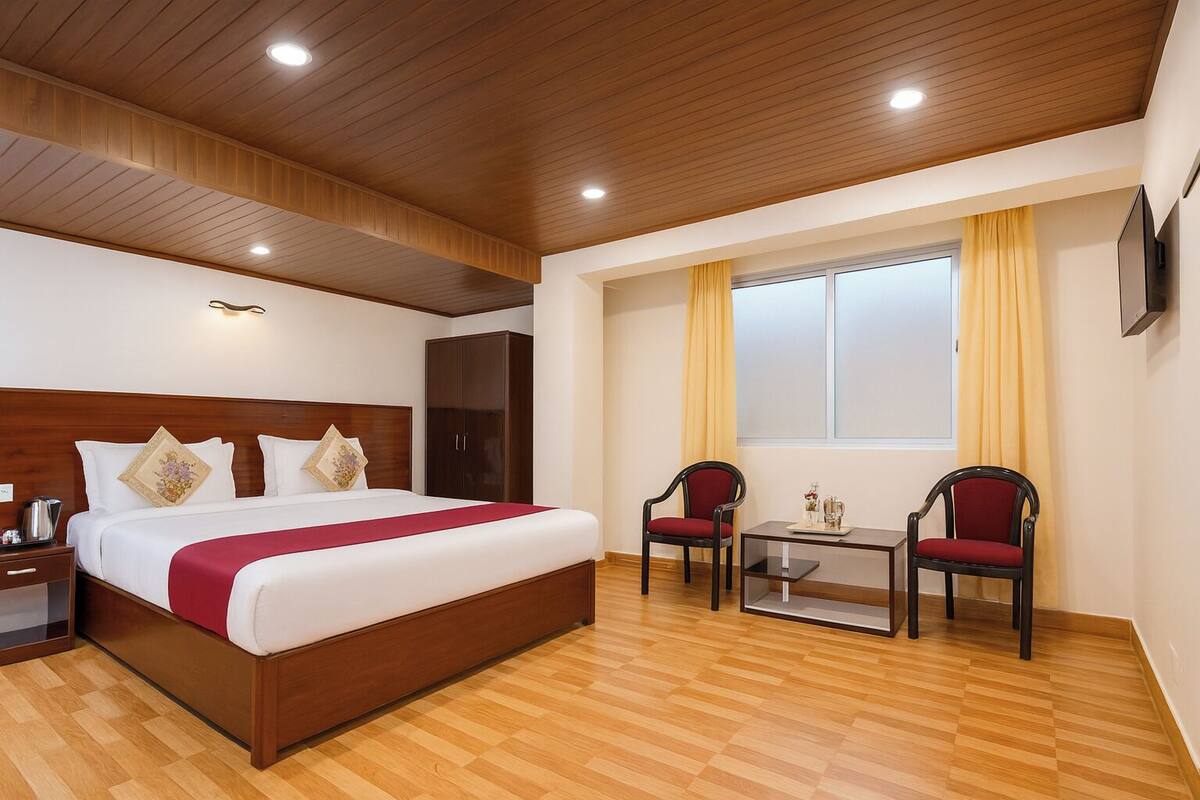 Quarto de hotel com teto de madeira, uma cama grande, um guarda-roupa, uma escrivaninha com duas cadeiras e uma janela com cortinas.