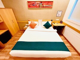 Quarto de hotel com cama, mesa de cabeceira, escrivaninha, cadeira, guarda-roupa e janela.