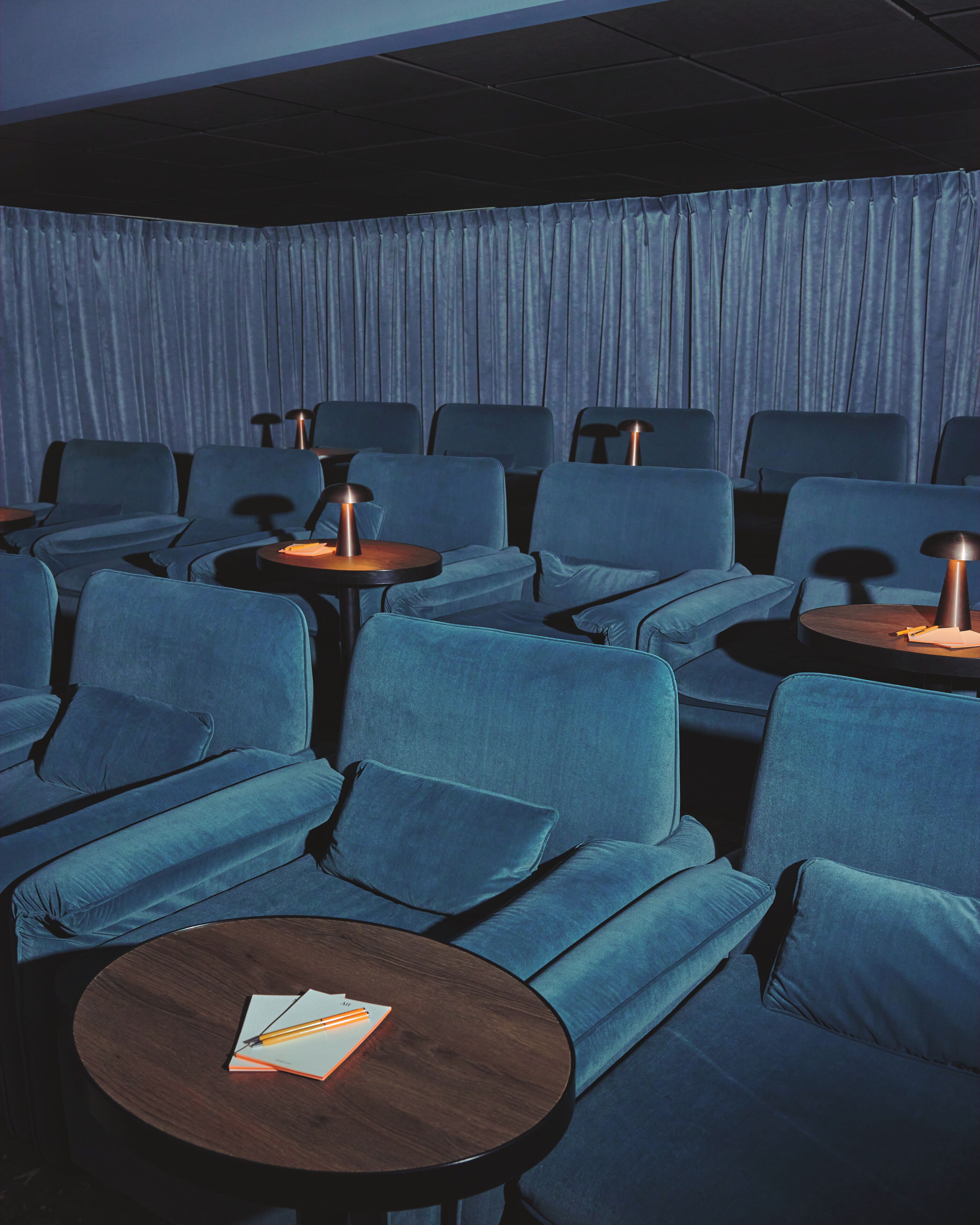 Um cinema com fileiras de assentos azuis, mesas de madeira e luminárias.