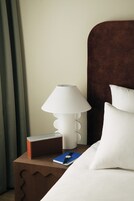 Uma cama bem arrumada com almofadas brancas, uma luminária branca sobre uma mesa de cabeceira de madeira e um livro ao lado da luminária.