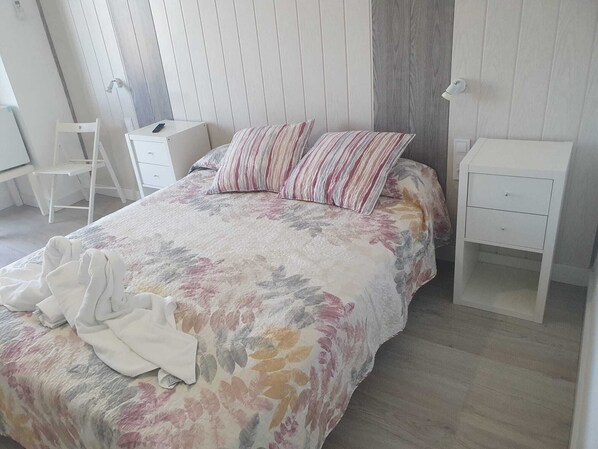 Quarto com colcha florida, duas almofadas listradas, mesa de cabeceira com gavetas, uma cadeira e escrivaninha.