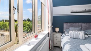 Quarto com uma janela ampla, uma cama com roupa de cama listrada, uma mesa de cabeceira com abajur e vista para o verde lá fora.