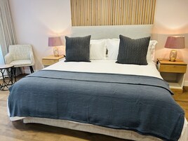 Cama bem arrumada com manta azul escura, duas luminárias de apoio e uma cadeira em quarto com piso de madeira e parede listrada.