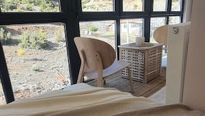 Um quarto com grandes janelas, uma cadeira de madeira, uma mesinha e uma cama.