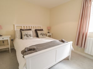 Quarto com cama branca, duas mesas de cabeceira, abajur e janela com cortinas.
