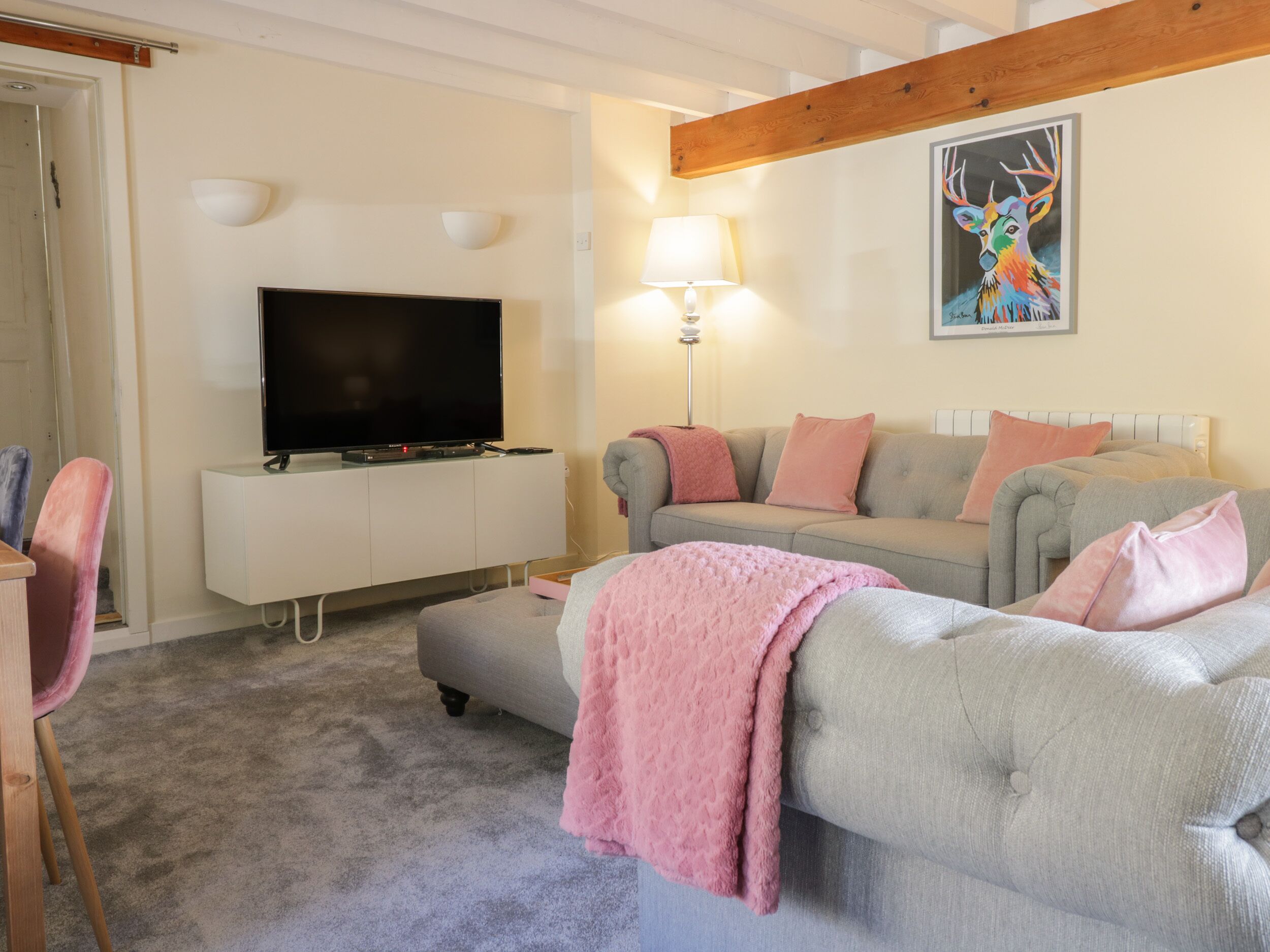 Uma sala de estar com uma TV de tela plana sobre um armário branco, um sofá cinza com almofadas rosa, um cobertor rosa e um quadro de um veado na parede.