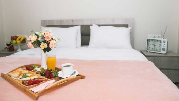 Uma cama bem arrumada com lençóis brancos, uma mesa de cabeceira com um relógio e uma bandeja com itens de café da manhã, incluindo torradas, ovos e um copo de suco de laranja.