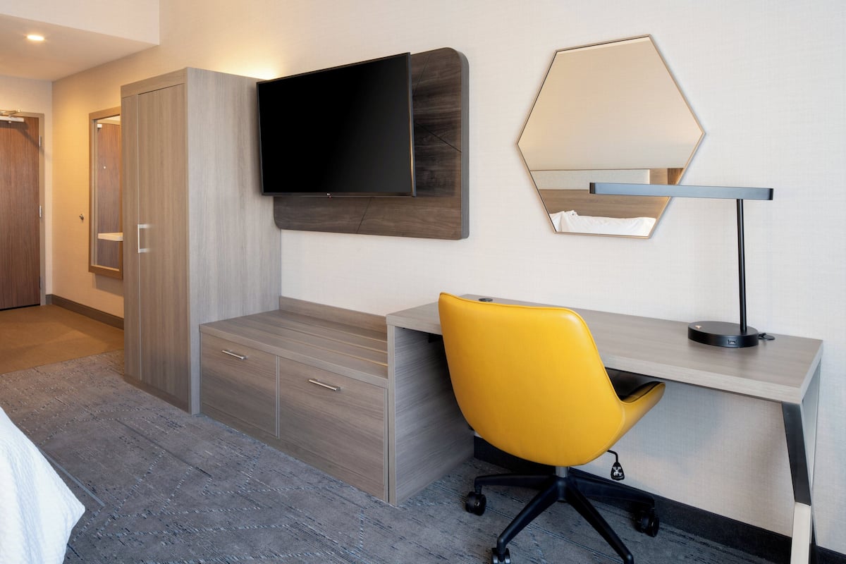 Quarto de hotel moderno com armário de madeira, TV de tela plana, escrivaninha com cadeira amarela e luminária na parede.