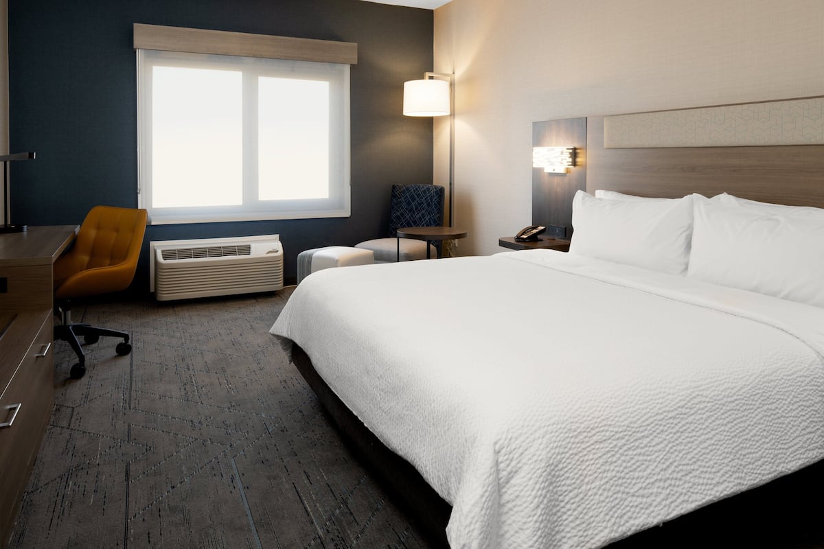 Quarto de hotel com uma cama grande, uma escrivaninha com cadeira, uma mesinha pequena, um abajur e uma janela com persianas.