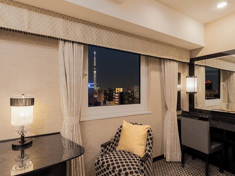 Quarto de hotel com vista para a cidade, uma mesa com luminária, uma cadeira com almofada e um espelho grande.