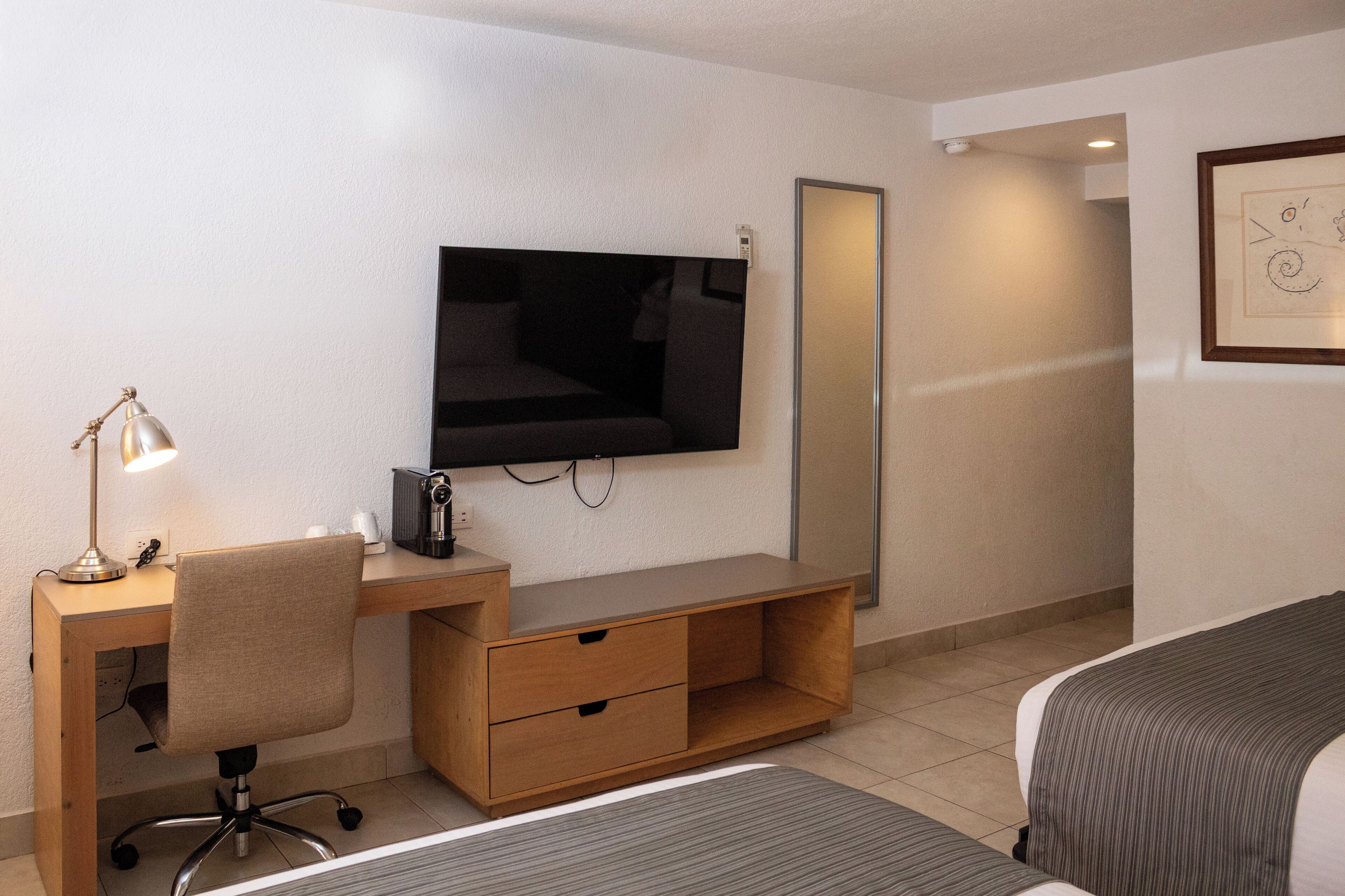 Quarto de hotel com escrivaninha de madeira, uma cadeira, televisão na parede, espelho e cama com colcha listrada.