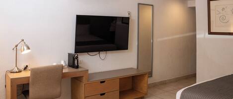 Quarto de hotel com escrivaninha de madeira, uma cadeira, televisão na parede, espelho e cama com colcha listrada.