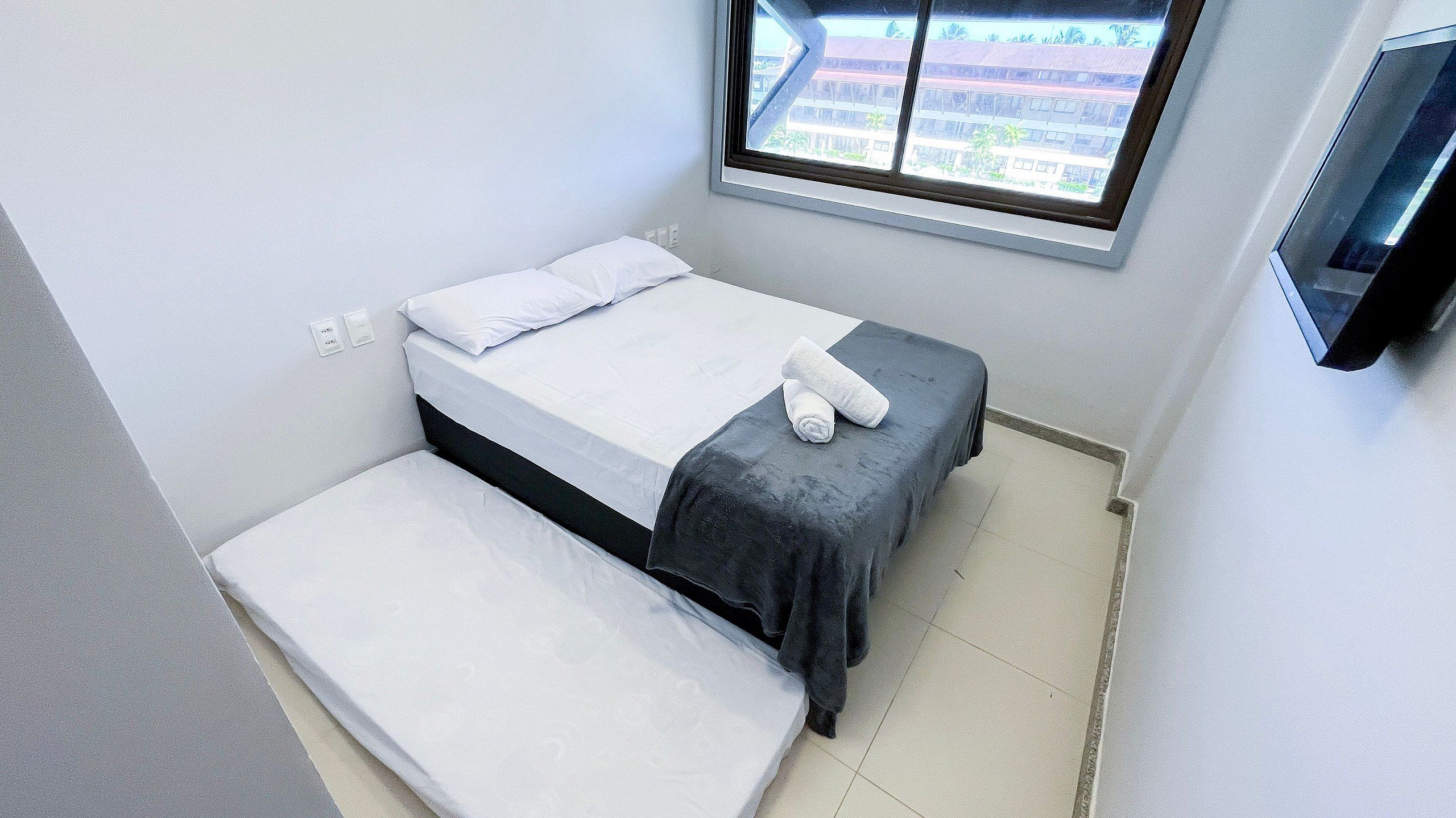 Uma cama de solteiro com colchão branco e manta escura, uma pequena mesa de cabeceira e uma janela com vista para edifícios.