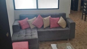 Um sofá modular cinzento com almofadas rosa e bege, um banquinho vermelho e uma mesa de centro de vidro com blocos brancos e cinzentos.