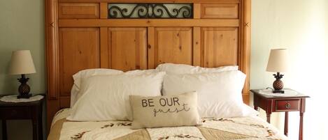 Cama bem arrumada com colcha, cabeceira de madeira, duas mesas de cabeceira com abajures e um travesseiro com a inscrição "BE OUR guest".