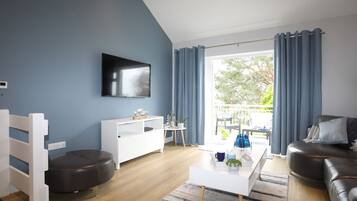 Uma sala de estar moderna com uma TV fixada na parede, um sofá branco, uma mesa de centro com objetos decorativos e vista para o exterior através de janelas amplas.