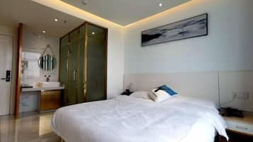 Quarto de hotel moderno com cama grande, mesa de cabeceira, luminária e uma obra de arte na parede. Banheiro com pia e espelho.