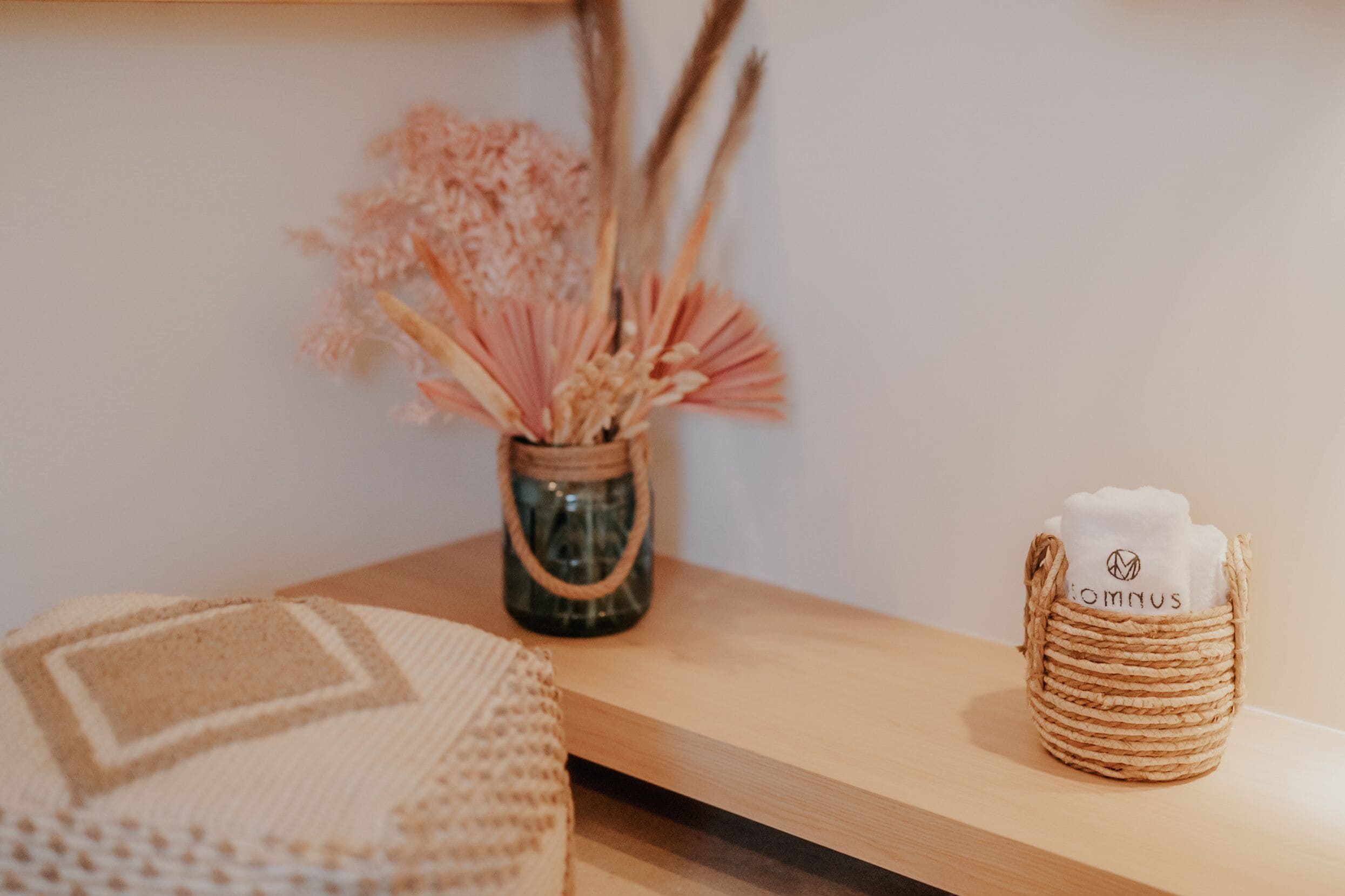 Uma mesa de madeira com uma cesta de palha contendo um pano branco e um pequeno vaso com flores secas.