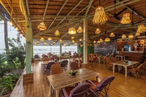 Restaurante ao ar livre com mesas e cadeiras de madeira, luminárias de vime penduradas e vista para o oceano.