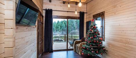 Interior em madeira com árvore de Natal, luminária pendente e vista por uma porta de vidro.
