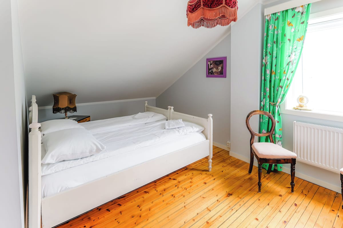 Quarto com cama branca, piso de madeira, uma cadeira, janela com cortinas verdes e radiador.