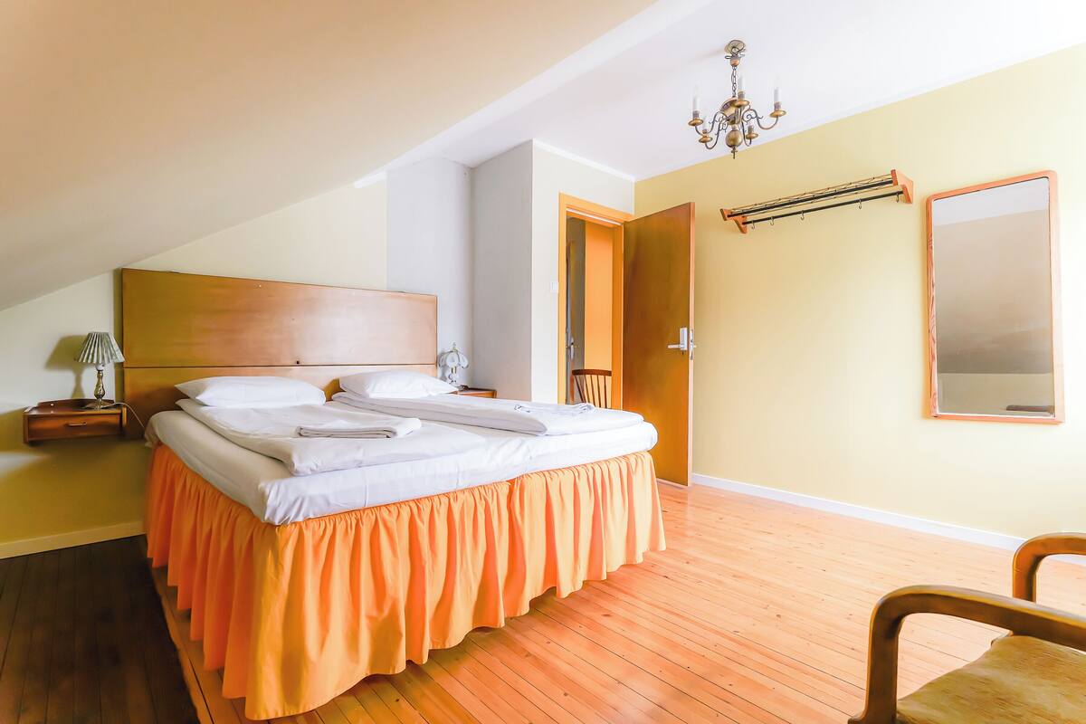 Quarto com cabeceira de madeira, cama com roupa de cama branca, colcha laranja, piso de madeira, mesa de cabeceira com abajur, cadeira, espelho e lustre.