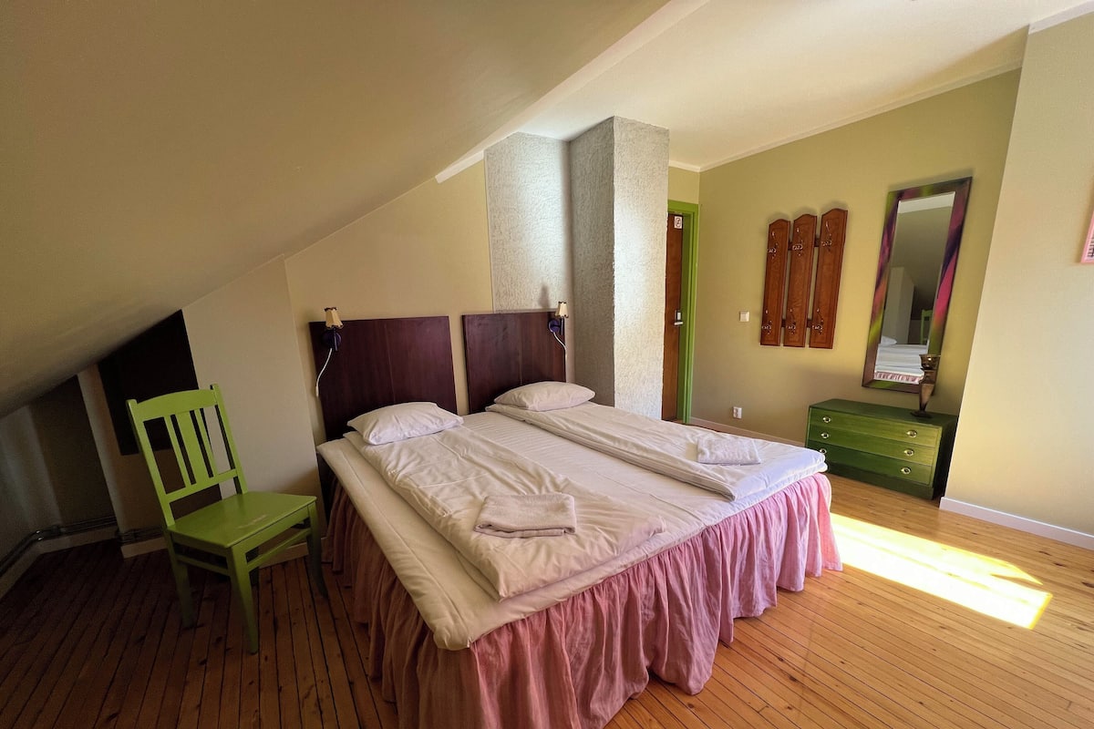 Quarto com uma cama grande, piso de madeira, uma cadeira verde, cômoda e espelho.