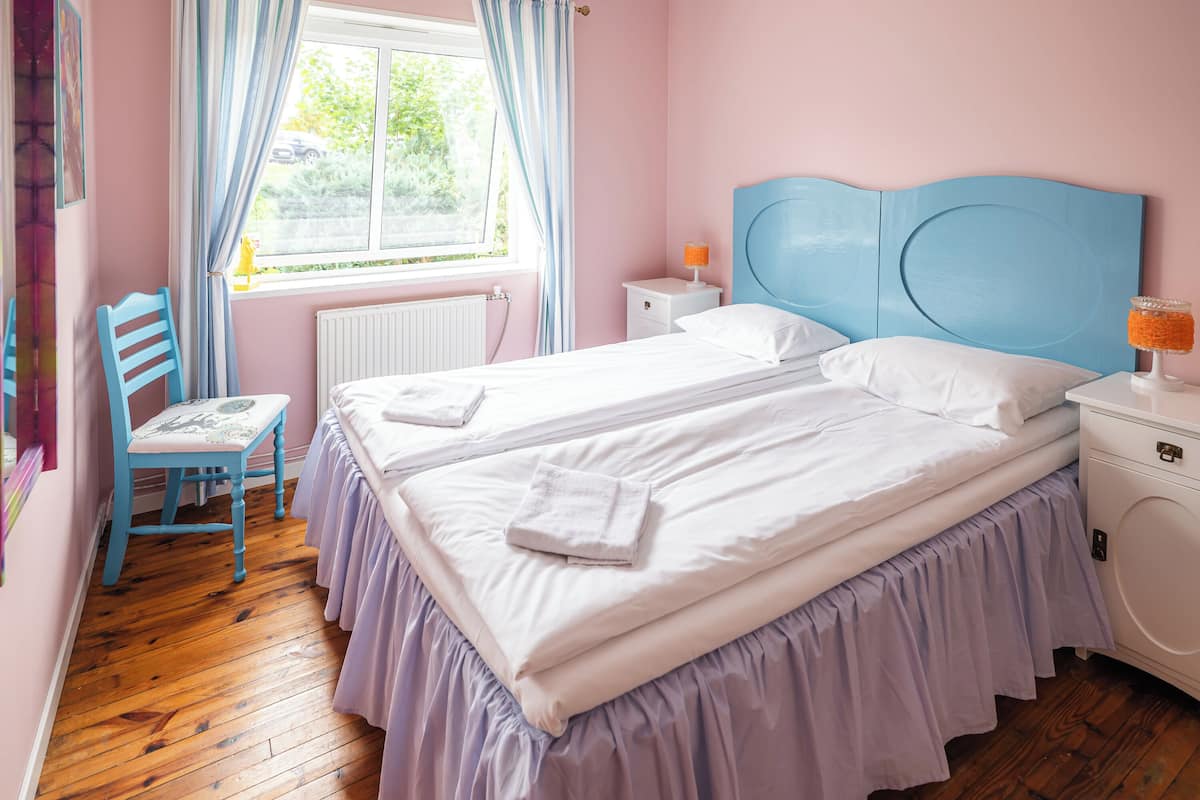 Quarto com duas camas, uma cadeira, uma mesa de cabeceira e uma janela com cortinas.
