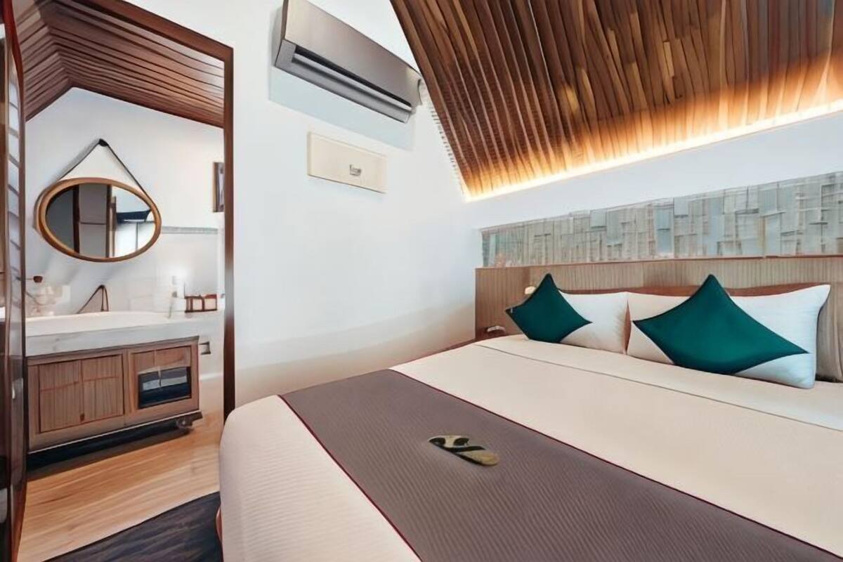 Quarto de hotel moderno com cama, cabeceira de madeira, espelho redondo, ar condicionado integrado e teto com design em ripas.