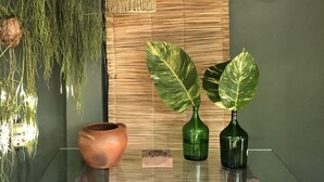 Uma mesa com tampo de vidro, dois vasos verdes e um cachepot de terracota, um abajur de fibra natural e plantas pendentes em frente a uma parede verde.