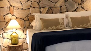 Quarto com parede de pedra, cama com roupa de cama branca e escura, e mesa de cabeceira com luminária.