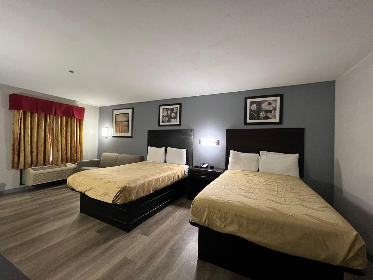 Quarto de hotel com duas camas, uma mesa de cabeceira, um abajur, uma janela com cortinas e um quadro na parede.