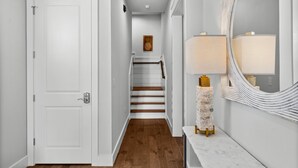 Um corredor com porta branca, escadas de madeira, um aparador branco e um espelho com moldura singular.