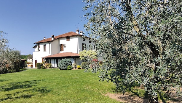 Uma casa de dois andares com telhado de telhas vermelhas, rodeada por um gramado bem cuidado e oliveiras.