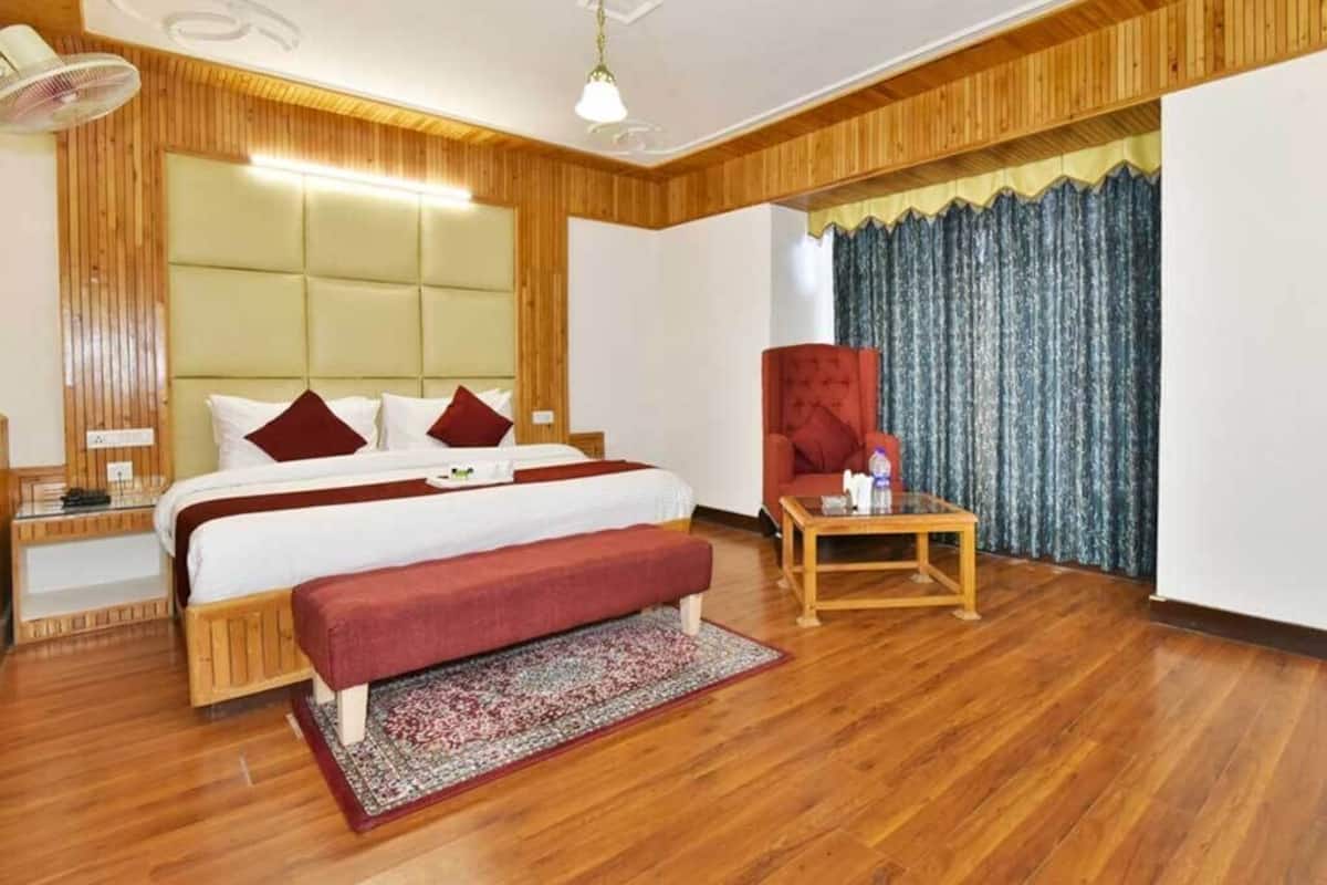 Quarto de hotel com piso de madeira, uma cama grande com roupa de cama vermelha e branca, uma poltrona vermelha, uma mesa pequena e uma janela com cortinas.