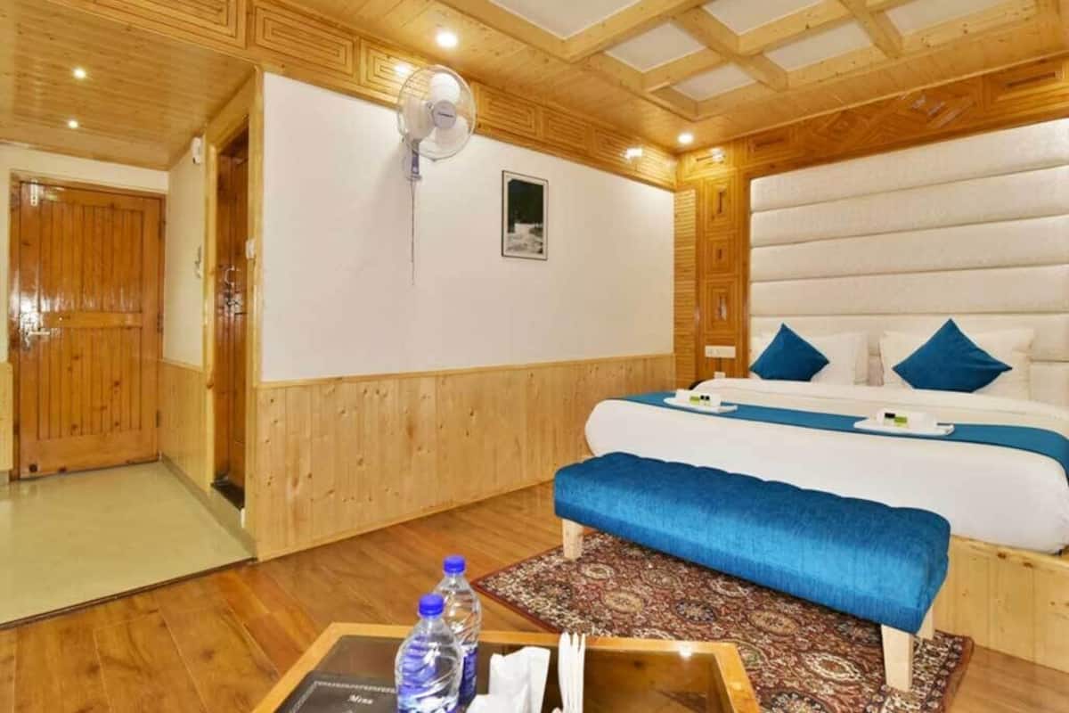 Quarto com painéis de madeira, uma cama, um ventilador e uma pequena mesa com uma garrafa de água.